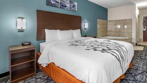 Quarto de hotel com uma cama grande, cabeceira, obra de arte na parede, mesa de cabeceira, banheiro com banheira e piso coberto por carpete.