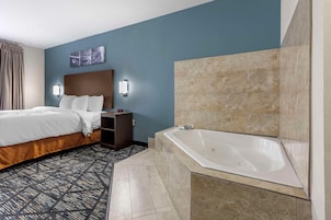 Quarto de hotel com cama, mesa de cabeceira, luminária na parede, um quadro e banheiro com banheira.