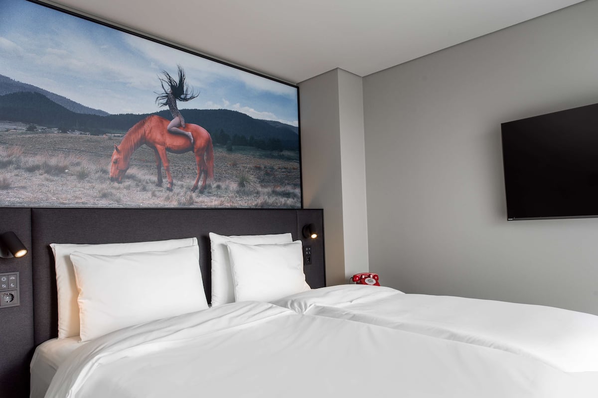 Quarto de hotel com uma cama grande, televisão montada e uma pintura de um cavalo numa montanha.