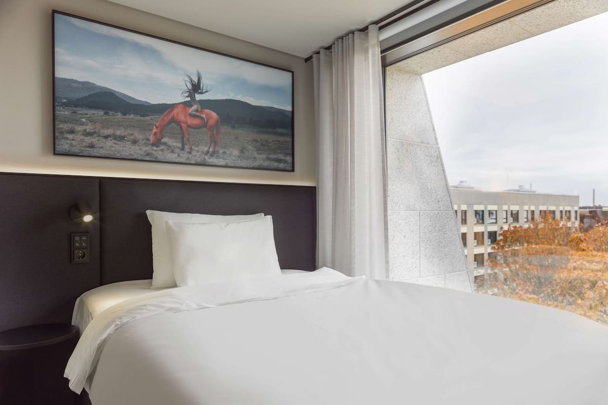 Quarto de hotel com uma cama, cabeceira, painel de controle e uma grande janela com vista para um edifício e árvores.