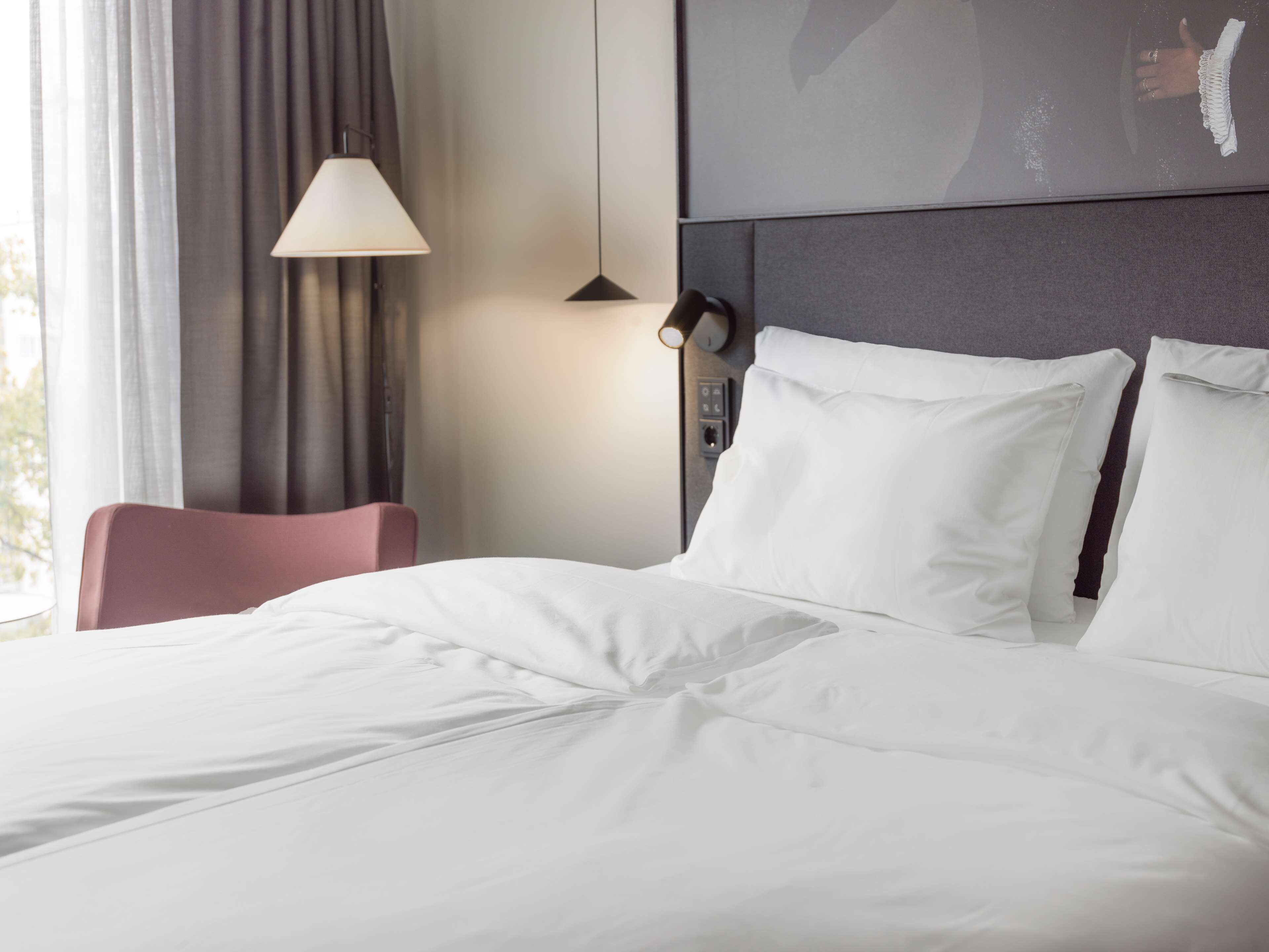Quarto de hotel com cama, cadeira, luminária e janela com cortinas.