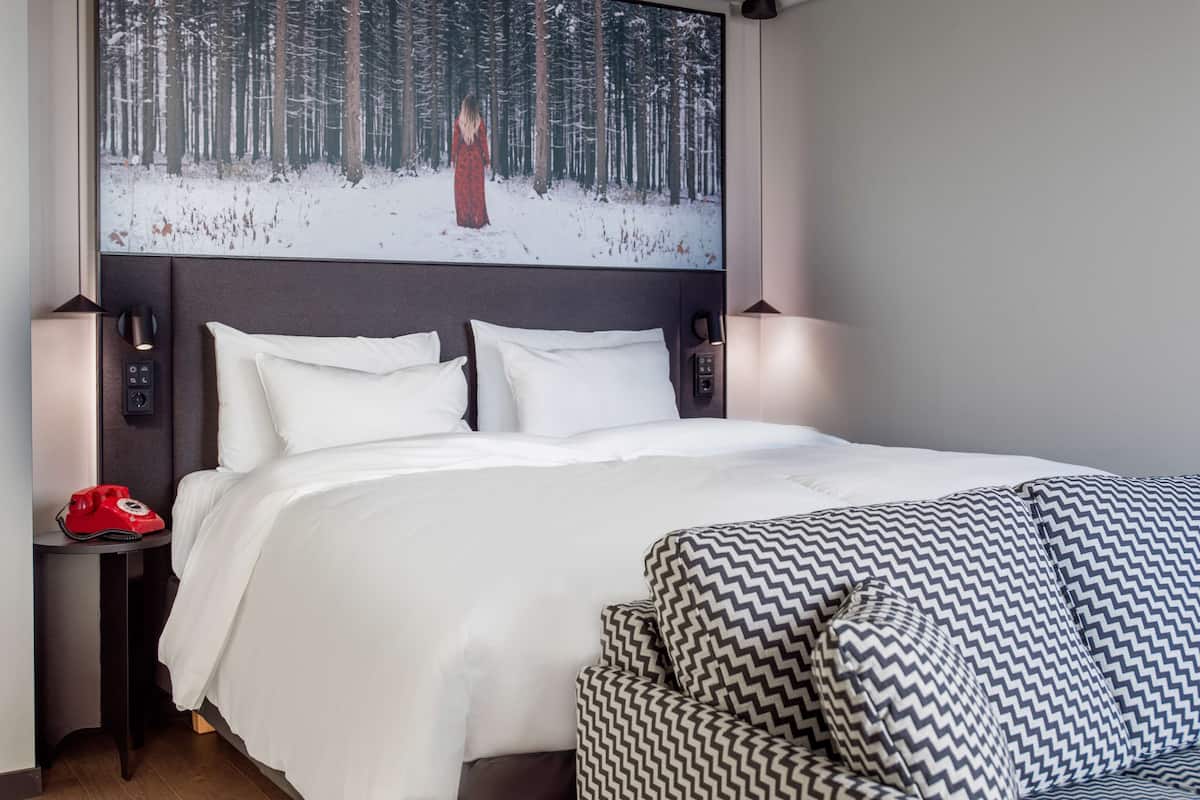 Quarto de hotel moderno com uma cama grande, cabeceira com arte temática de floresta, luminárias de cabeceira, telefone giratório vermelho e poltrona estampada.