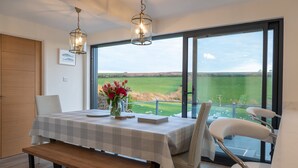 Uma área de refeições com mesa posta para dois, um banco e vista para um campo verde através de grandes janelas.