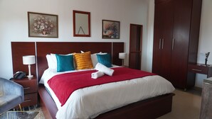 Cama bem arrumada com um cobertor vermelho, várias almofadas e uma toalha branca. Há quadros com flores na parede, um guarda-roupa de madeira e uma mesa com abajur.