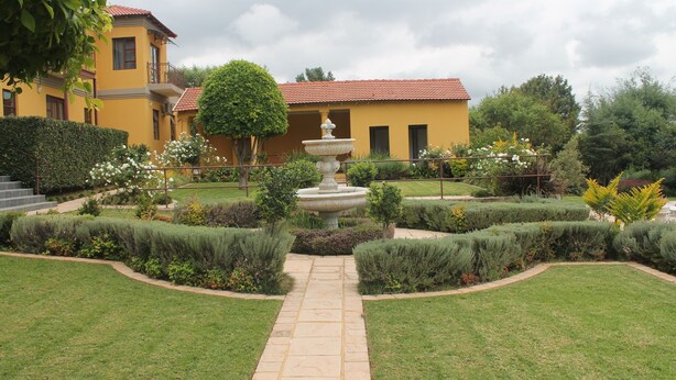 Um jardim bem cuidado com uma fonte central, sebes podadas e um caminho que leva a um edifício amarelo com telhado vermelho.