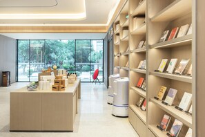 Uma biblioteca moderna com janelas amplas, uma mesa central com livros e caixas, e estantes repletas de livros.