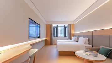 Quarto de hotel com cama, escrivaninha, cadeira, mesa redonda com chaleira e janela com vista para a cidade.