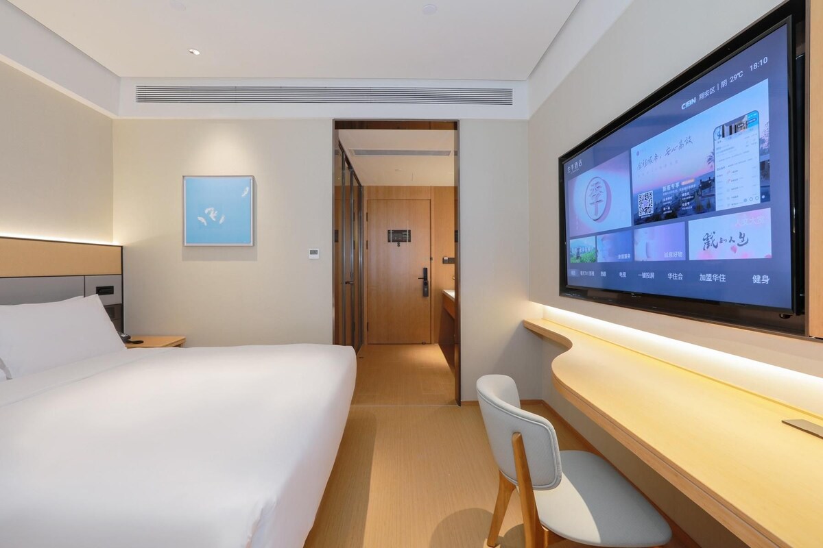 Quarto de hotel moderno com uma cama grande, uma mesa com cadeira, televisão na parede e porta para outro cômodo.