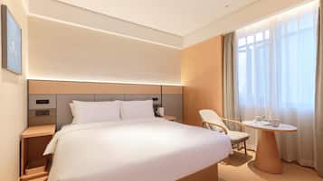Quarto de hotel moderno com uma cama grande, mesas de cabeceira, uma cadeira, uma pequena mesa redonda e uma janela com cortinas.