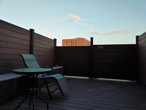 Um deck de madeira com uma mesa e duas cadeiras, cerca de madeira e um edifício ao fundo.
