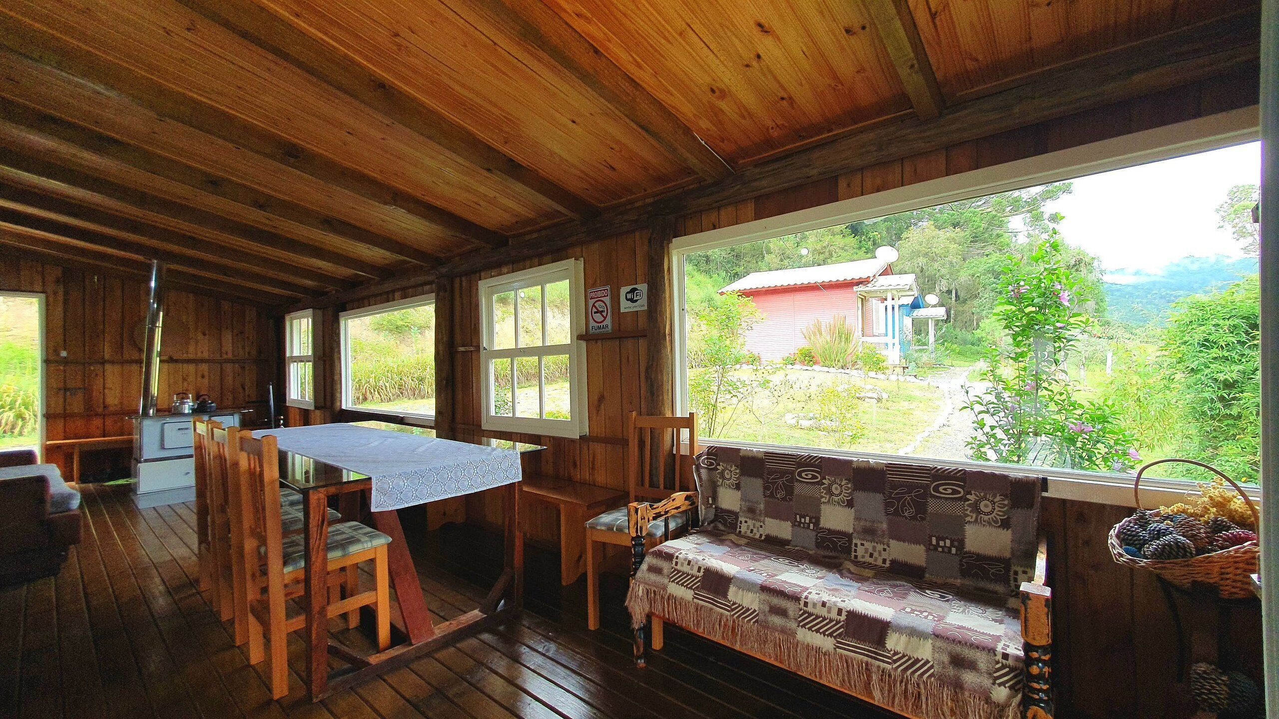 Uma cabana de madeira com mesa de jantar, cadeiras e sofá. Há janelas com vista para o verde e um edifício com telhado vermelho do lado de fora.