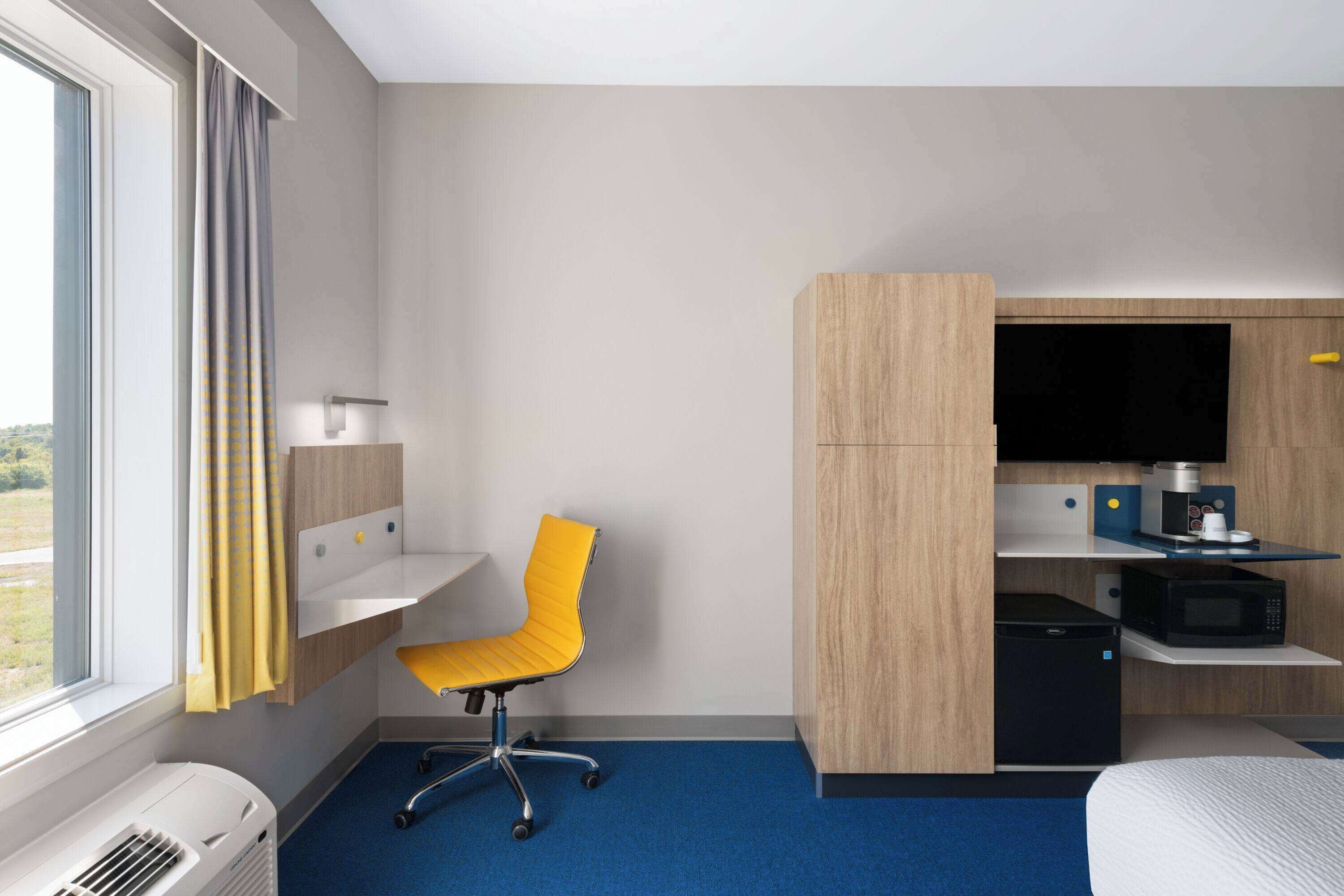 Quarto de hotel com escrivaninha, cadeira, cozinha compacta e uma grande janela.