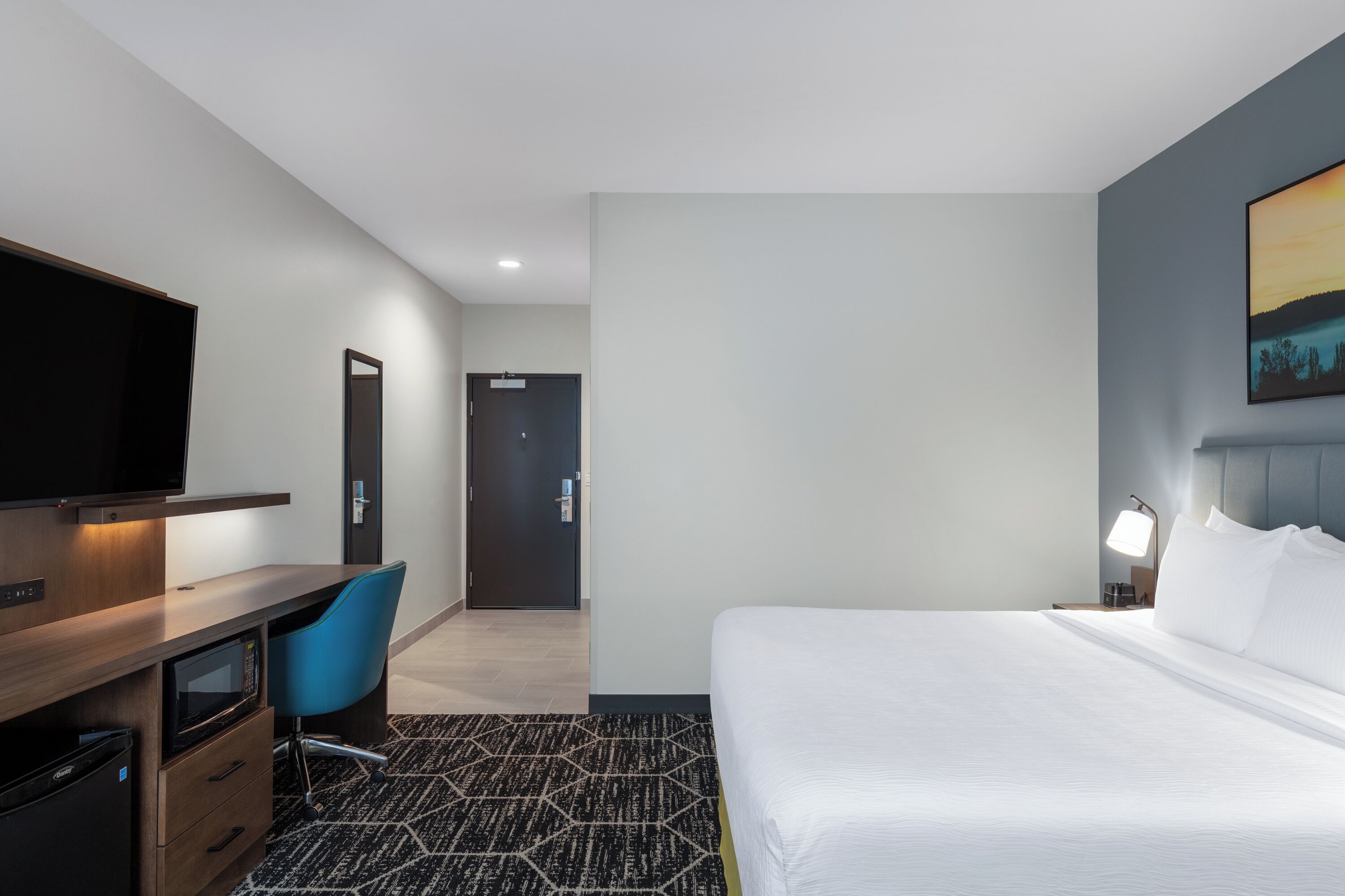 Quarto de hotel com cama, escrivaninha com computador, televisão e porta para outro cômodo.
