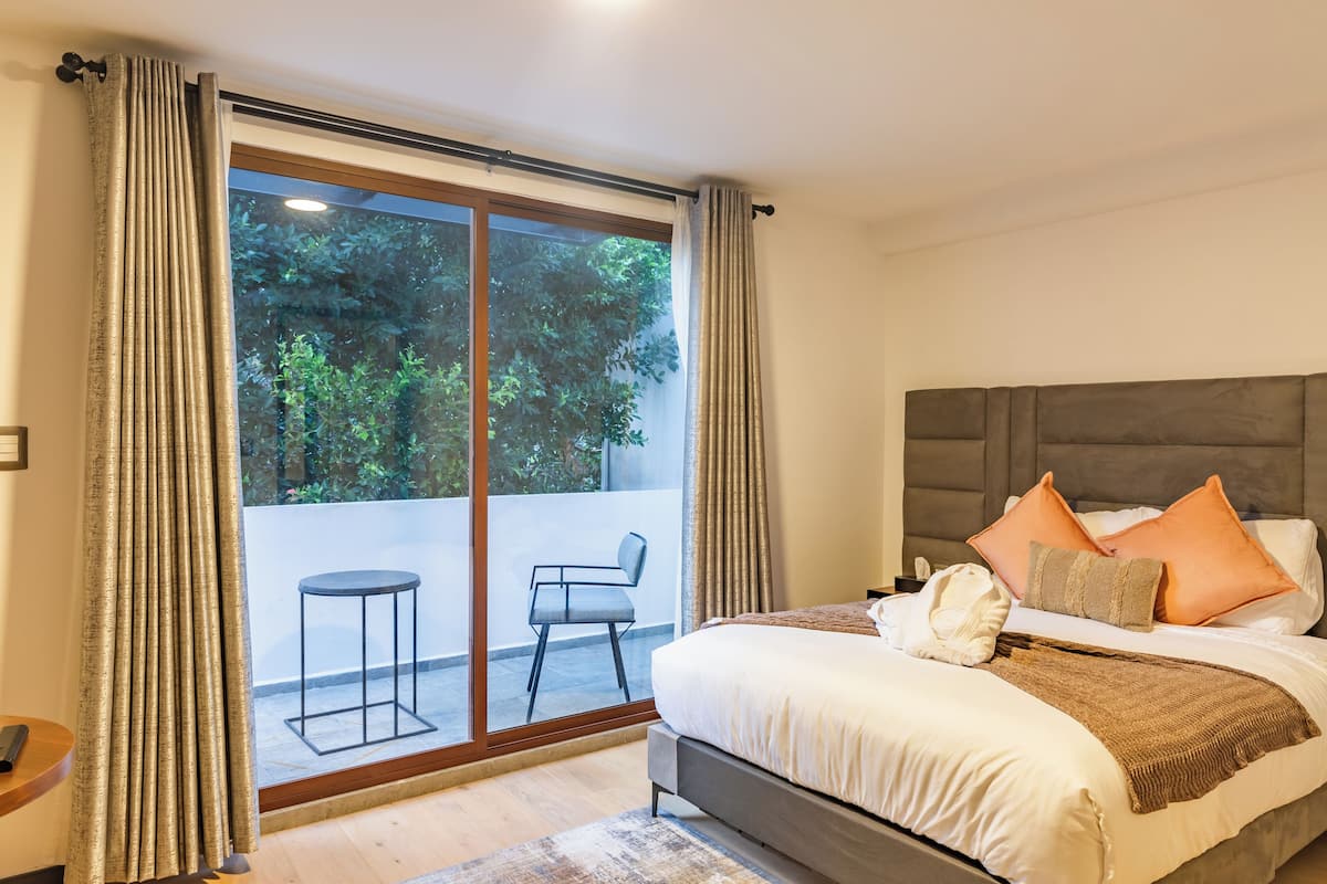 Quarto com cama grande, cabeceira e vista para uma varanda com cadeira e mesa.