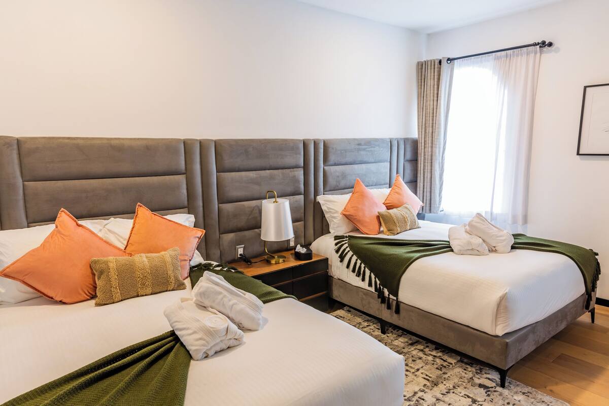Quarto de hotel com duas camas, mesa de cabeceira, luminária e janela com cortinas.
