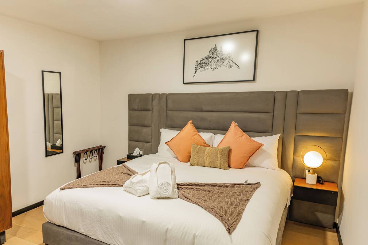 Cama bem arrumada com cabeceira, mesa de cabeceira com abajur e tapete com padrão geométrico.