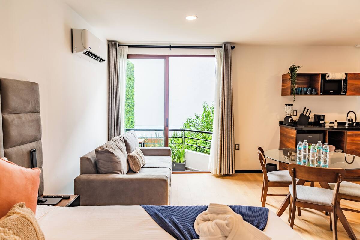 Quarto de hotel moderno com cama, sofá, mesa de jantar com cadeiras, cozinha e varanda com vista para o verde.