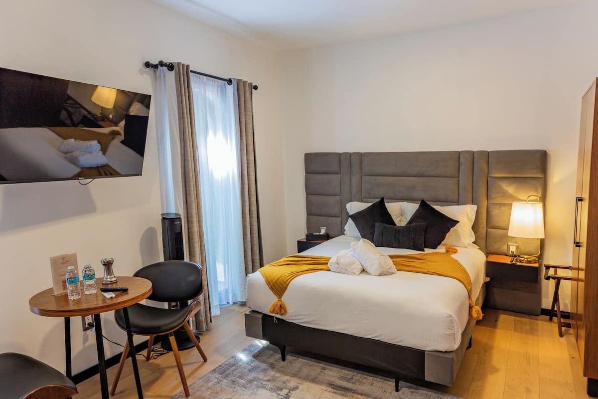 Quarto de hotel com cama, escrivaninha, cadeira, televisão e mesa de cabeceira com abajur.