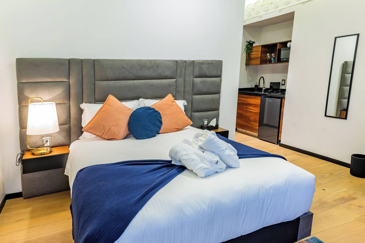 Quarto de hotel moderno com uma cama grande, uma mesinha de cabeceira com abajur, uma pequena escrivaninha e uma área de cozinha planejada.