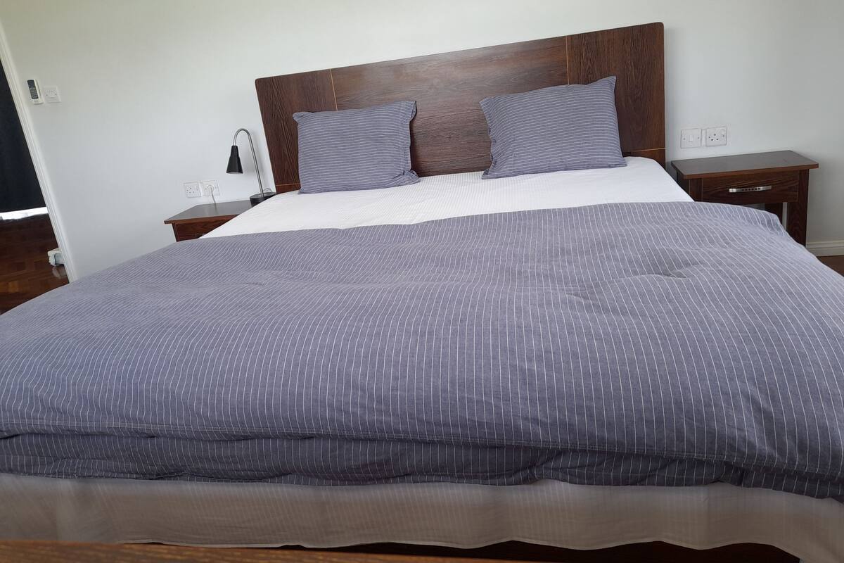 Cama bem arrumada com colcha listrada e duas almofadas, cabeceira de madeira, mesa de cabeceira com luminária e um criado-mudo com tomada.