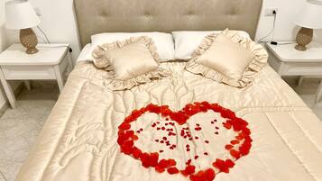 Cama arrumada com um arranjo de pétalas de rosa em formato de coração, ladeado por dois travesseiros com bordas franzidas.