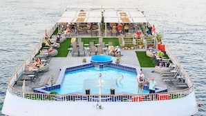 Um navio de cruzeiro com área de piscina, assentos externos e um convés coberto com mesas e cadeiras.