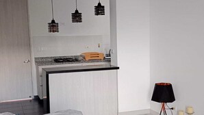 Uma sala de estar moderna com um sofá azul, uma mesa de centro branca e uma área de cozinha com bancada e luminárias pendentes.