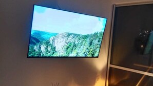 Televisão de tela plana presa na parede, um armário de madeira embaixo e uma janela com vista para uma paisagem de montanhas.