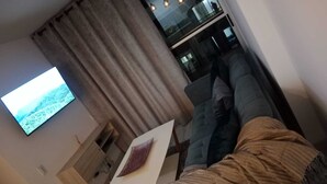 Uma sala de estar com televisão de tela plana na parede, um sofá, uma mesa de centro e uma pessoa sentada no sofá.
