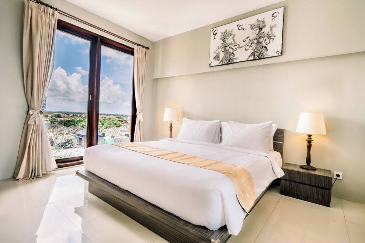 Quarto de hotel com uma cama grande, duas luminárias de cabeceira, vista da cidade pela janela e uma obra de arte na parede.