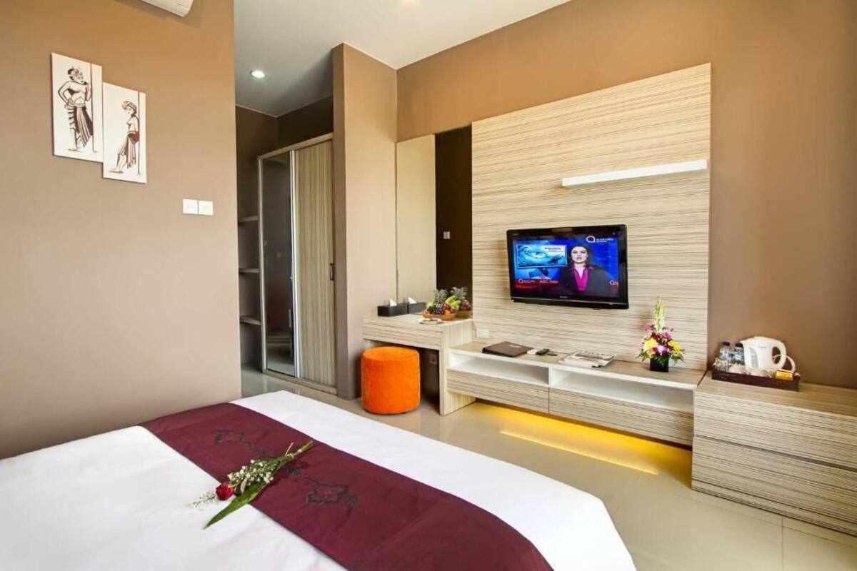 Quarto de hotel moderno com uma cama grande, uma TV fixada na parede, um banquinho laranja pequeno e uma mesa consola de madeira com um vaso de flores.