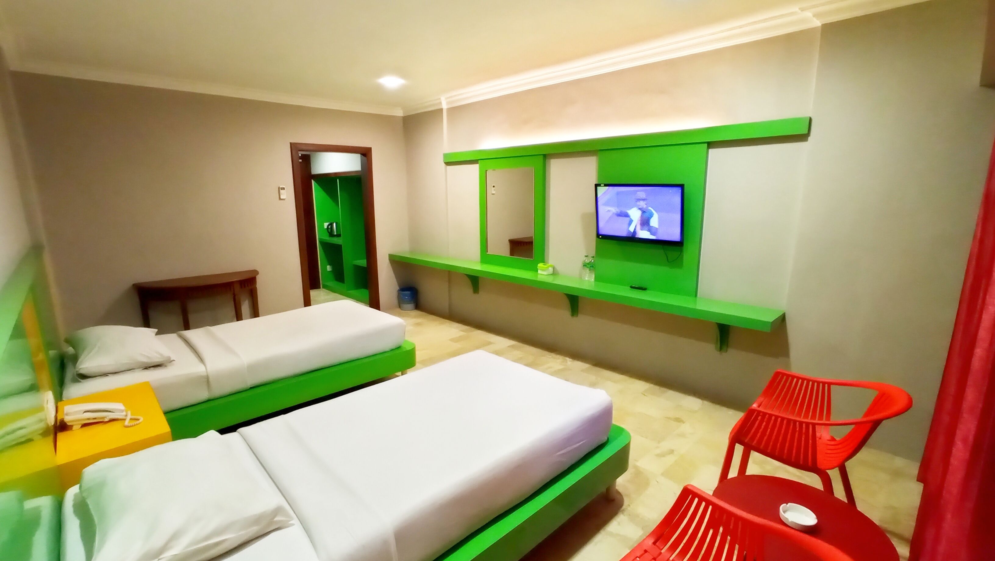 Quarto de hotel com duas camas, uma televisão presa a uma prateleira verde, uma mesinha com um telefone e uma cadeira vermelha.