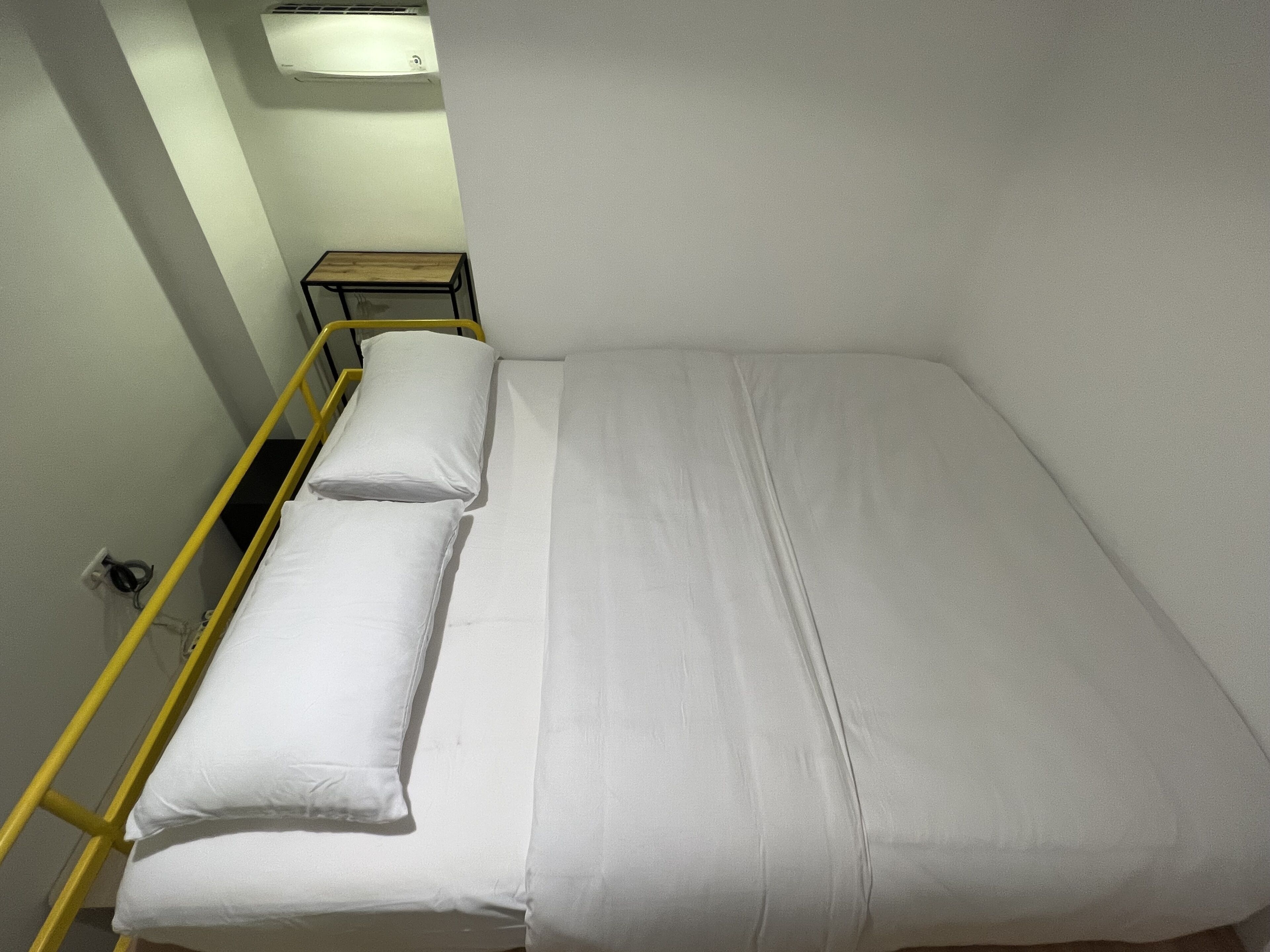 Uma cama de solteiro com roupa de cama branca em um quarto com uma escada amarela e uma pequena mesa com abajur.