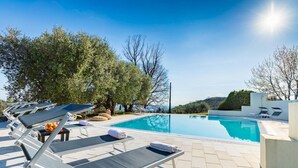 Uma área na beira da piscina com espreguiçadeiras e uma piscina azul cristalina, cercada por árvores e com vista para as colinas.