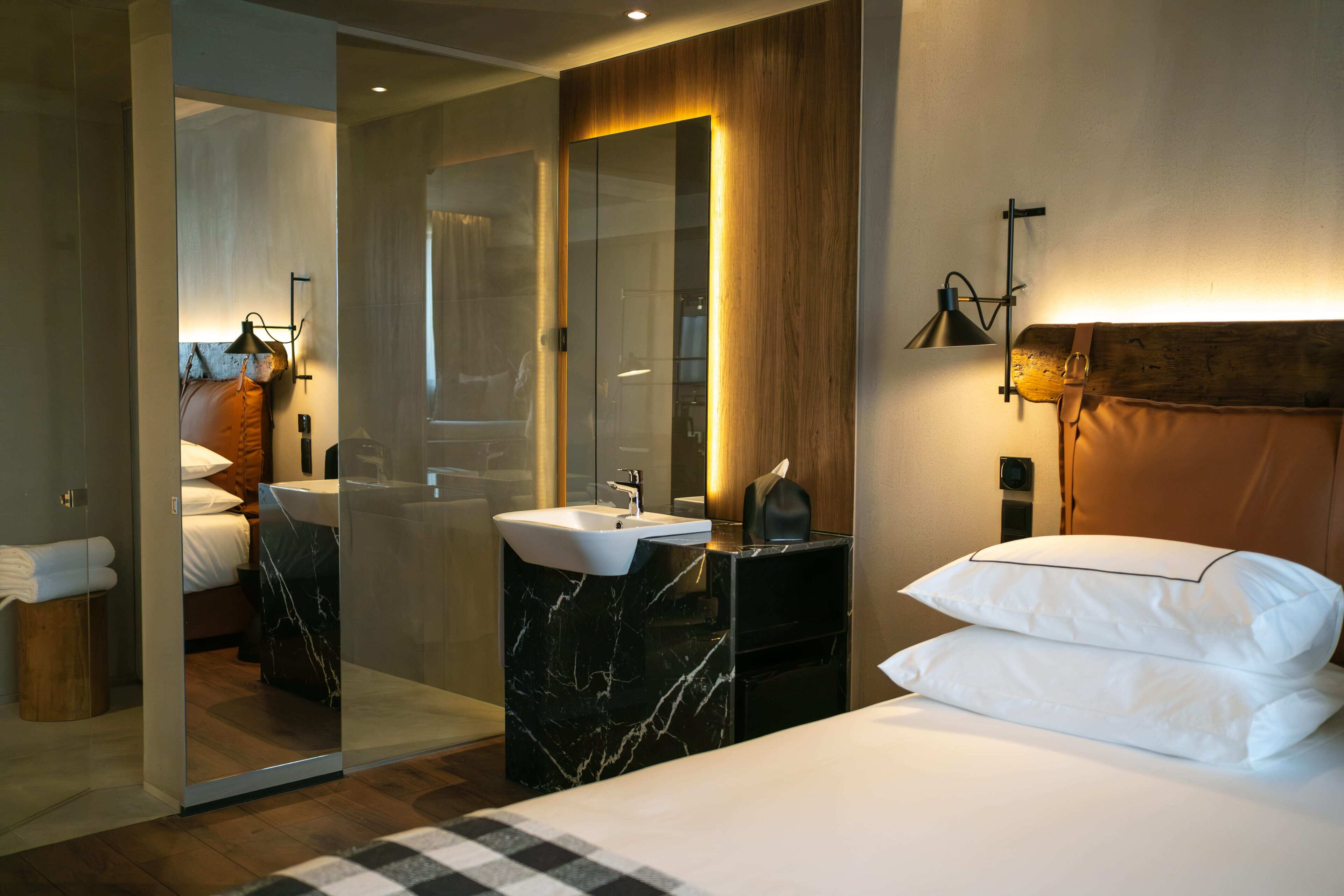 Quarto de hotel moderno com chuveiro em cabine de vidro, toucador de mármore, cama com roupa de cama branca e cabeceira de madeira.