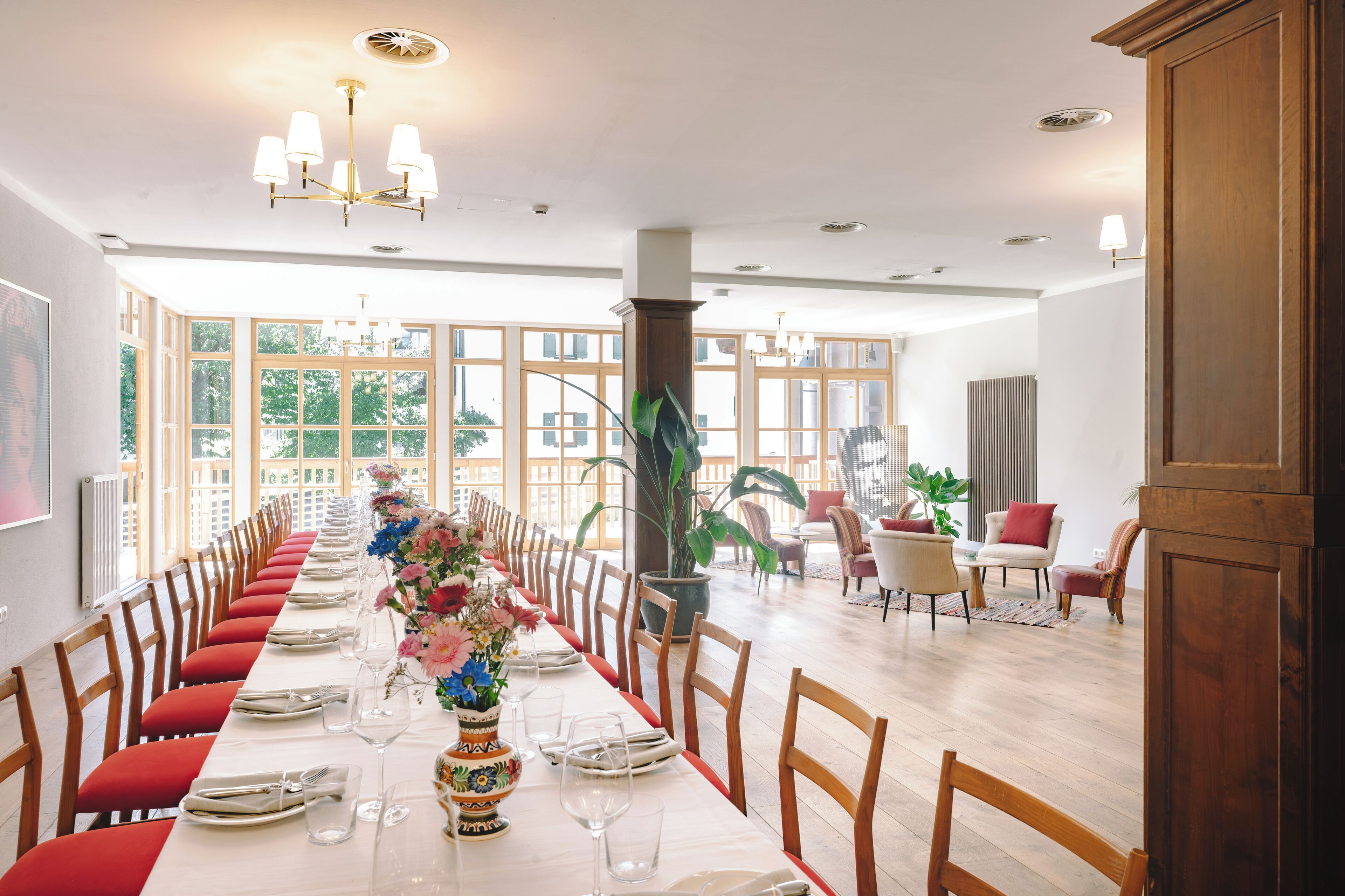 Uma mesa de jantar comprida posta para uma refeição em um espaço amplo com janelas grandes, cadeiras de madeira e um lustre.