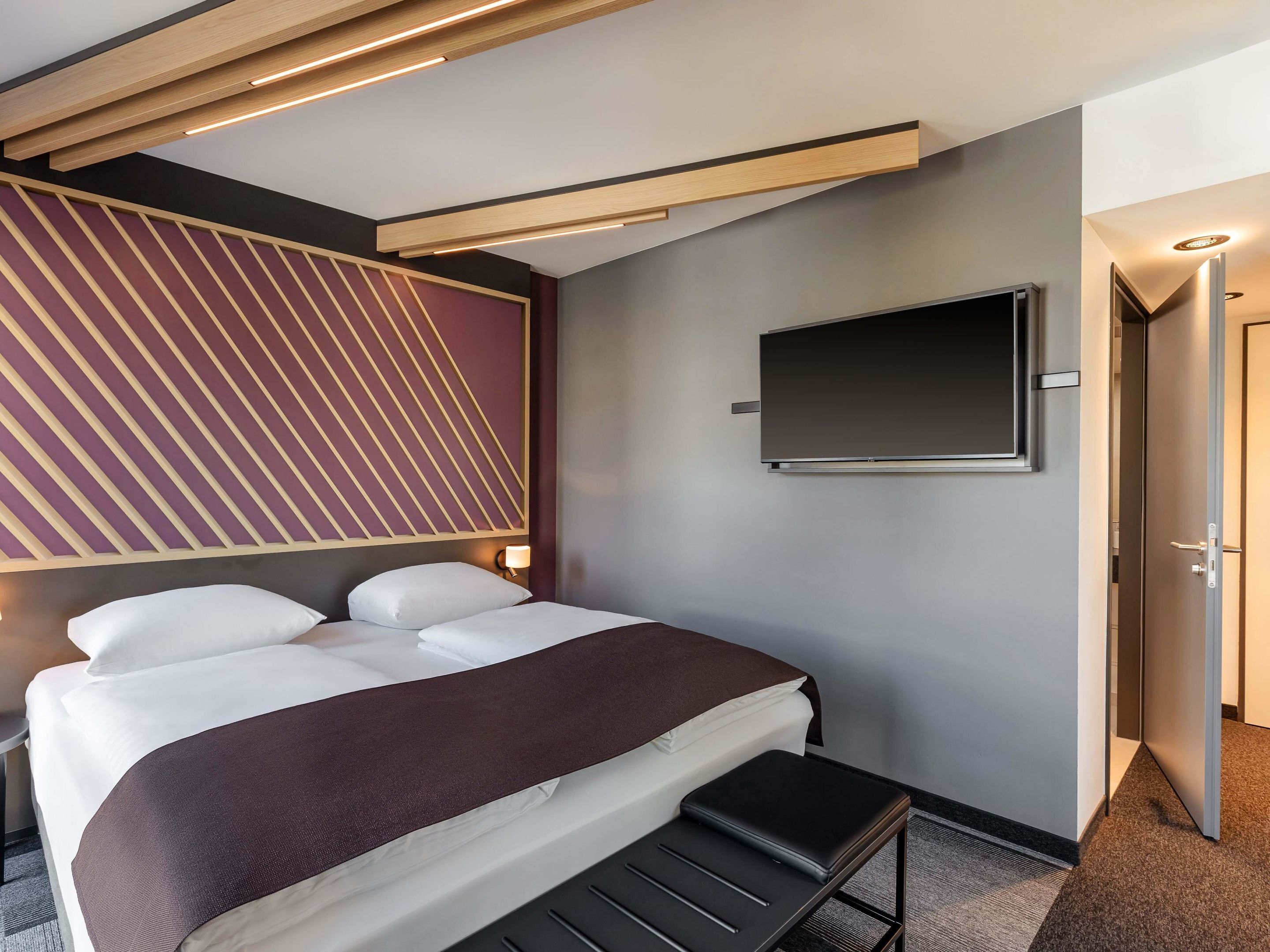 Quarto de hotel moderno com uma cama bem arrumada, uma televisão de tela plana na parede e uma porta que dá para outro quarto.