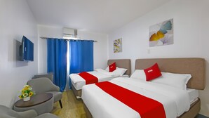 Quarto de hotel com duas camas, um sofá, uma mesinha com um vaso de flores e uma televisão na parede.