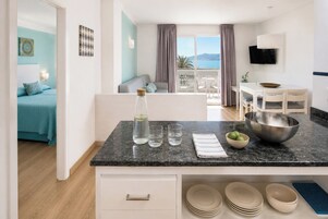 Uma cozinha moderna com bancada de mármore, uma área de jantar com mesa e cadeiras, e um quarto com cama e vista para o mar.