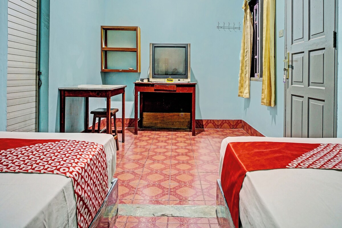 Um quarto pequeno com duas camas, uma escrivaninha com televisão e uma janela com cortinas.
