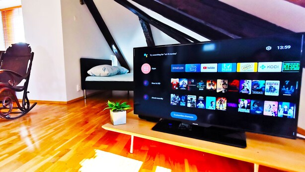 Uma sala de estar com piso de madeira, uma TV preta sobre um suporte, uma cadeira preta, uma pequena planta e uma escada ao fundo.