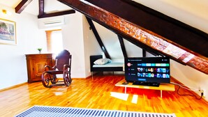 Um quarto com teto inclinado, piso de madeira, televisão em um suporte, uma poltrona de balanço e uma cama com travesseiro.