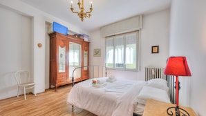 Quarto com piso de madeira, cama com roupa de cama branca, abajur vermelho, armário de madeira, uma cadeira e janela com cortinas de renda.