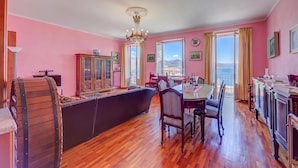 Quarto com piso de madeira, uma mesa de jantar com cadeiras, um sofá, um candelabro e janelas amplas com cortinas.