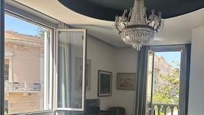 Quarto de hotel com uma grande janela, uma escrivaninha, uma cadeira, uma cama com colcha verde e um candelabro.