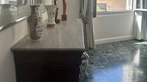 Quarto com mesa de mármore exibindo objetos decorativos, um armário de madeira escura e vista de um edifício através de uma janela com cortinas.