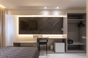 Quarto de hotel moderno com cama, escrivaninha, cadeira, televisão presa na parede, mini-geladeira e armário.