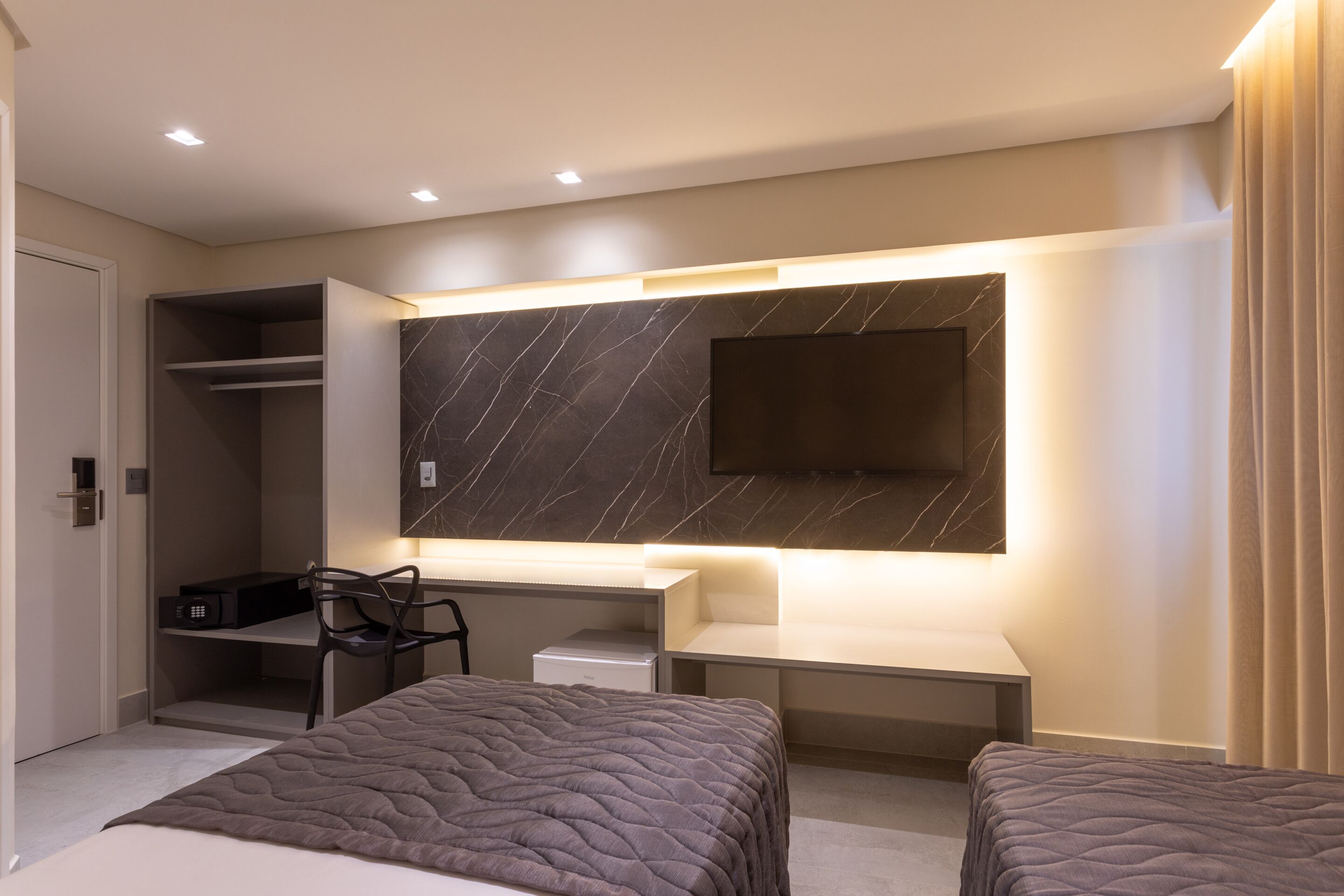 Quarto de hotel moderno com uma cama grande, uma escrivaninha, uma cadeira e televisão integrada.