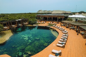 Um resort com piscina transparente, deck de madeira e construções com telhado de palha.