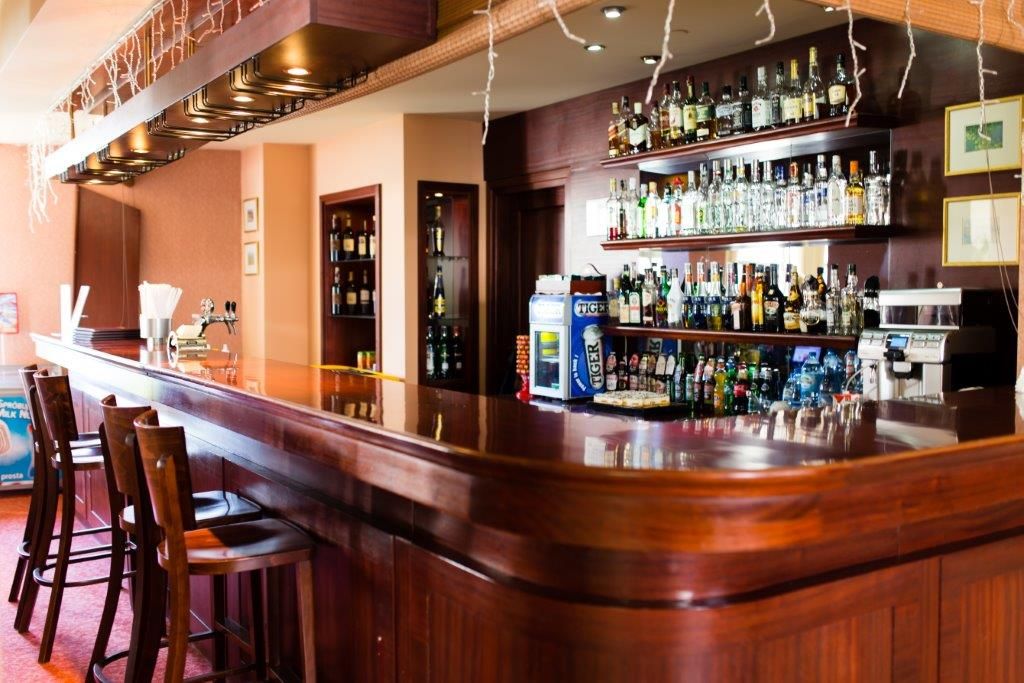 Uma área de bar com balcão e banquetas de madeira, prateleiras com garrafas de bebidas e um refrigerador de cerveja Tiger.
